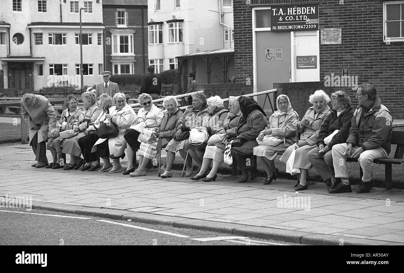 Large number of old people who are mostly women sitting on a long bench ...