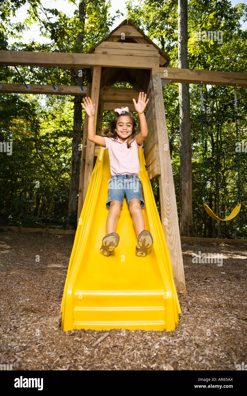 Hispanic girl sliding down outdoor slide with arms raised above head ...