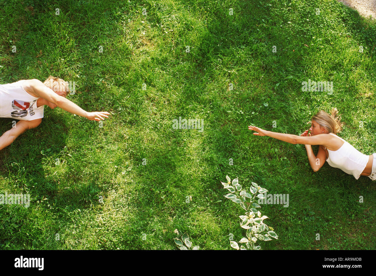 Young man and woman reaching for each other across sunlit green grass ...