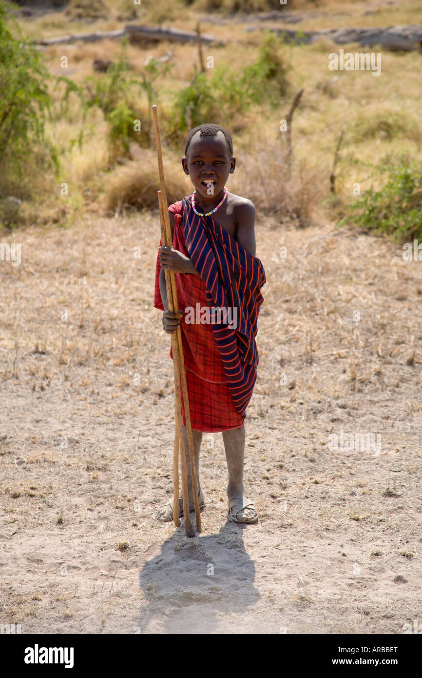 A young Maasai boy in traditional clothing holds several cattle sticks ...