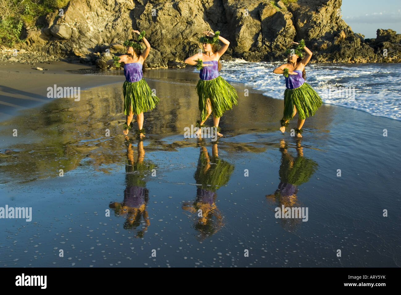 Three female hula dancers perform at the water's edge, their image is ...