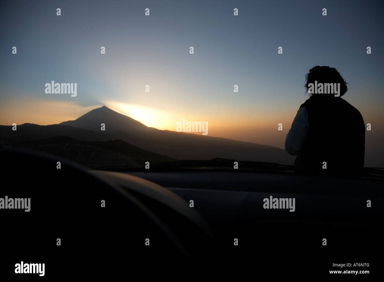 woman sitting on the bonnet of a car watching the sun setting over ...