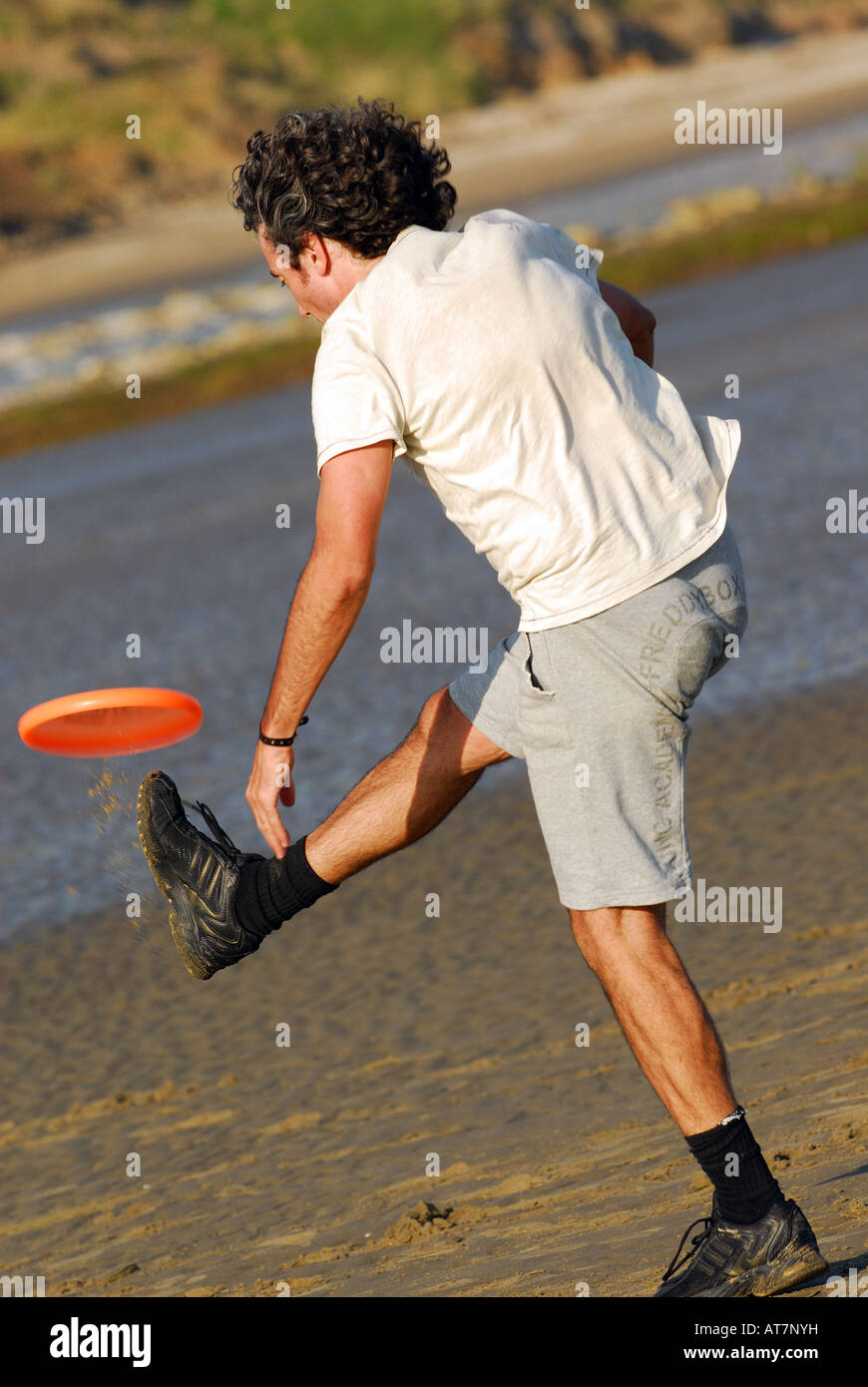 a young man playing with a frizbee on the beach kicking the disc and ...