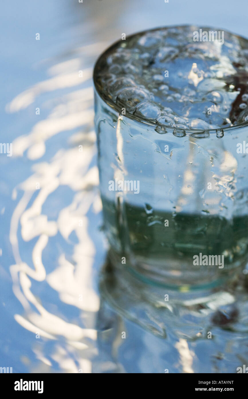Water pouring into a glass that stands on a mirror which reflects blue ...