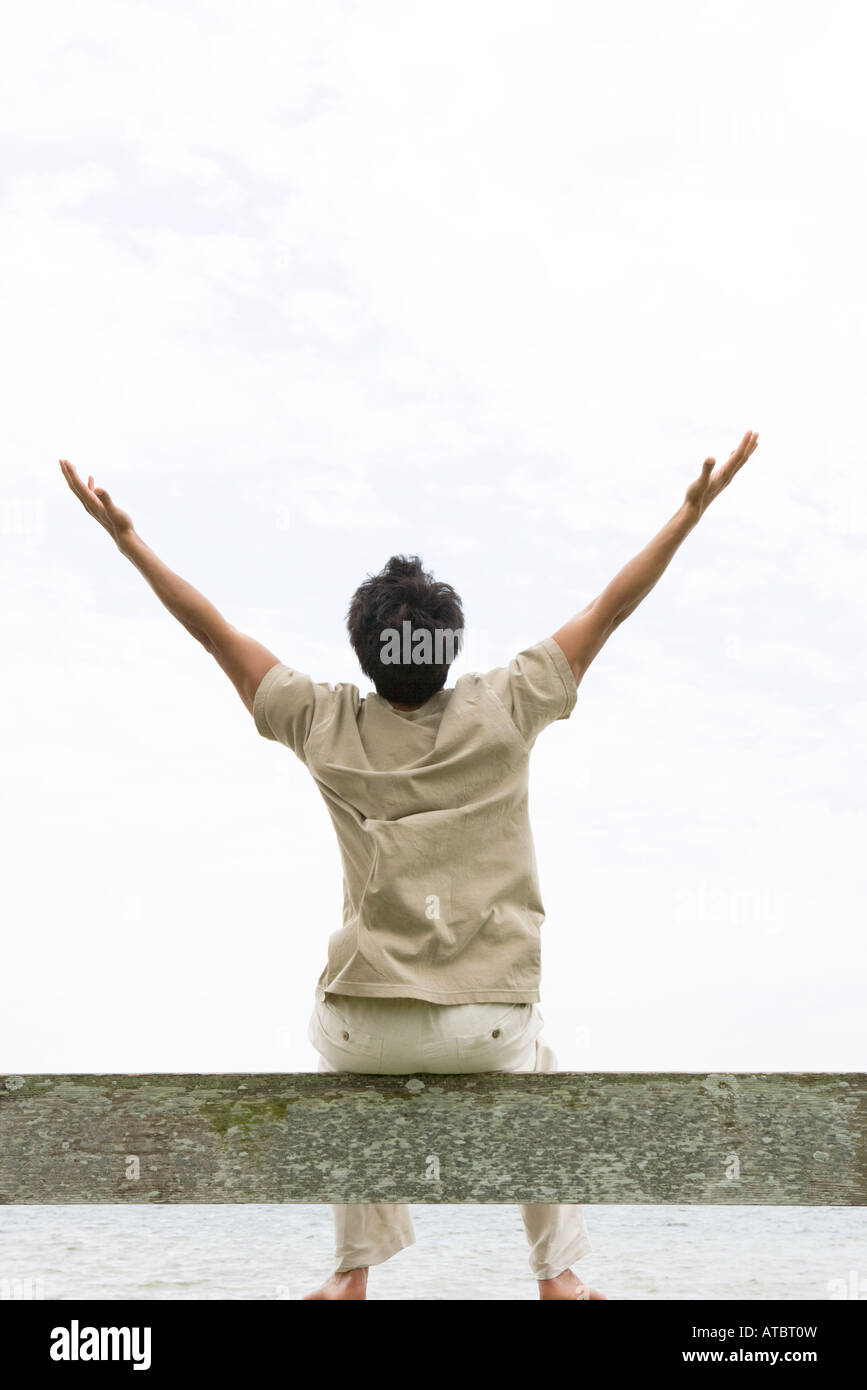 Man sitting on guard rail with arms raised, rear view Stock Photo - Alamy