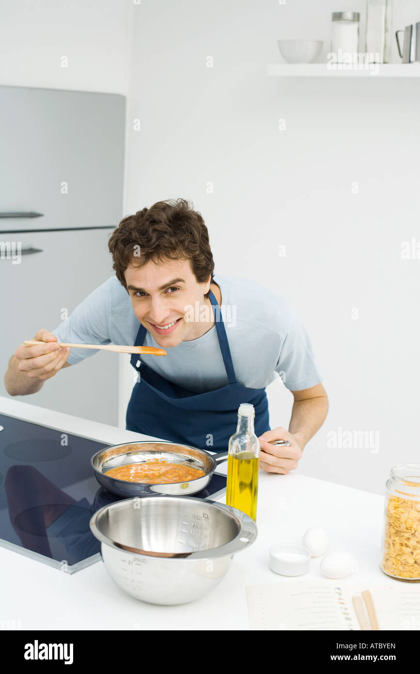 Man cooking in kitchen, bending over to taste sauce, smiling at camera ...