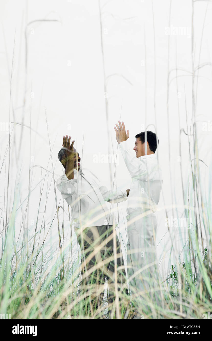 Two men greeting each other outdoors, hands raised, viewed through tall ...