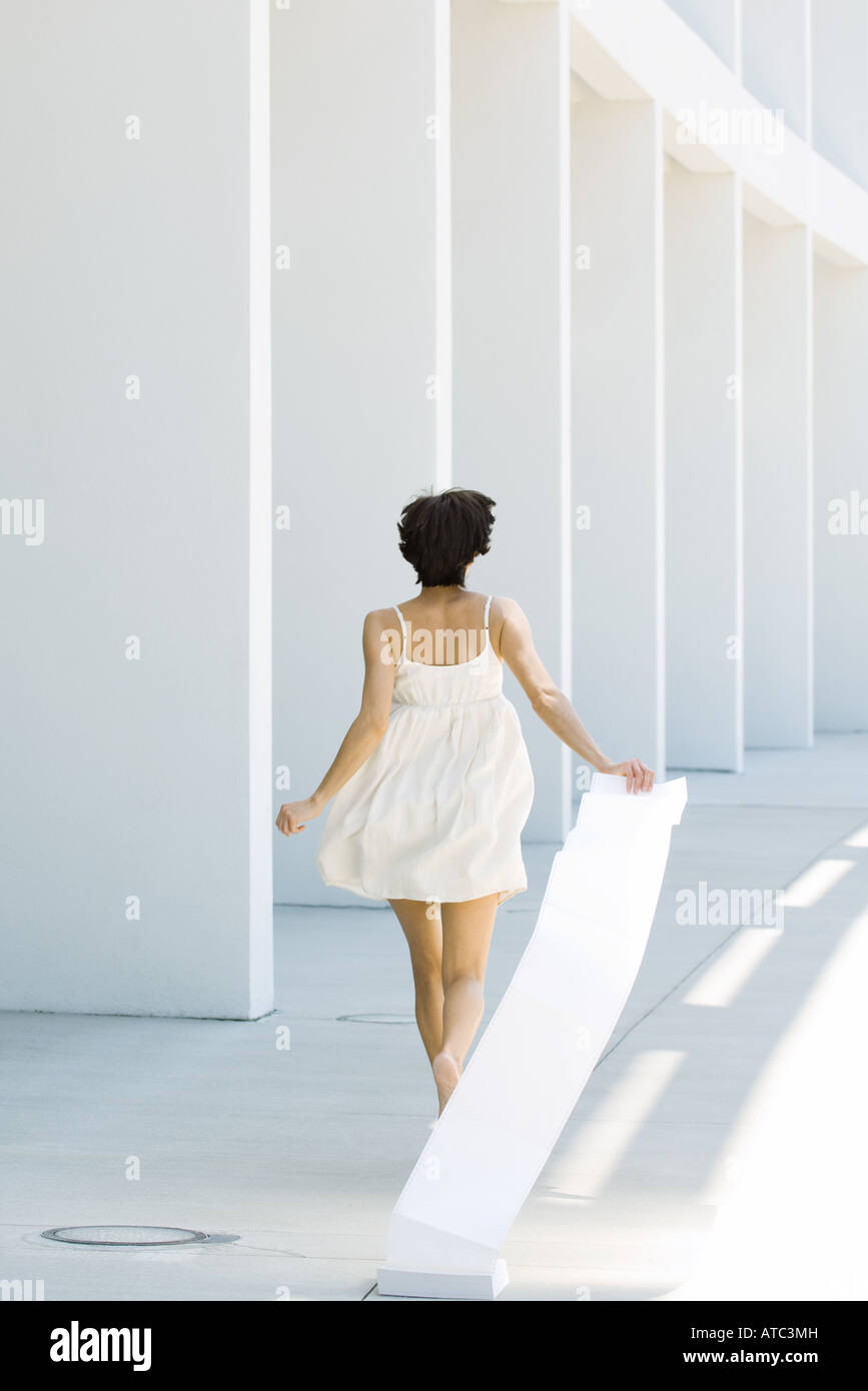 Woman in sundress running outdoors, pulling paper behind her, rear view ...