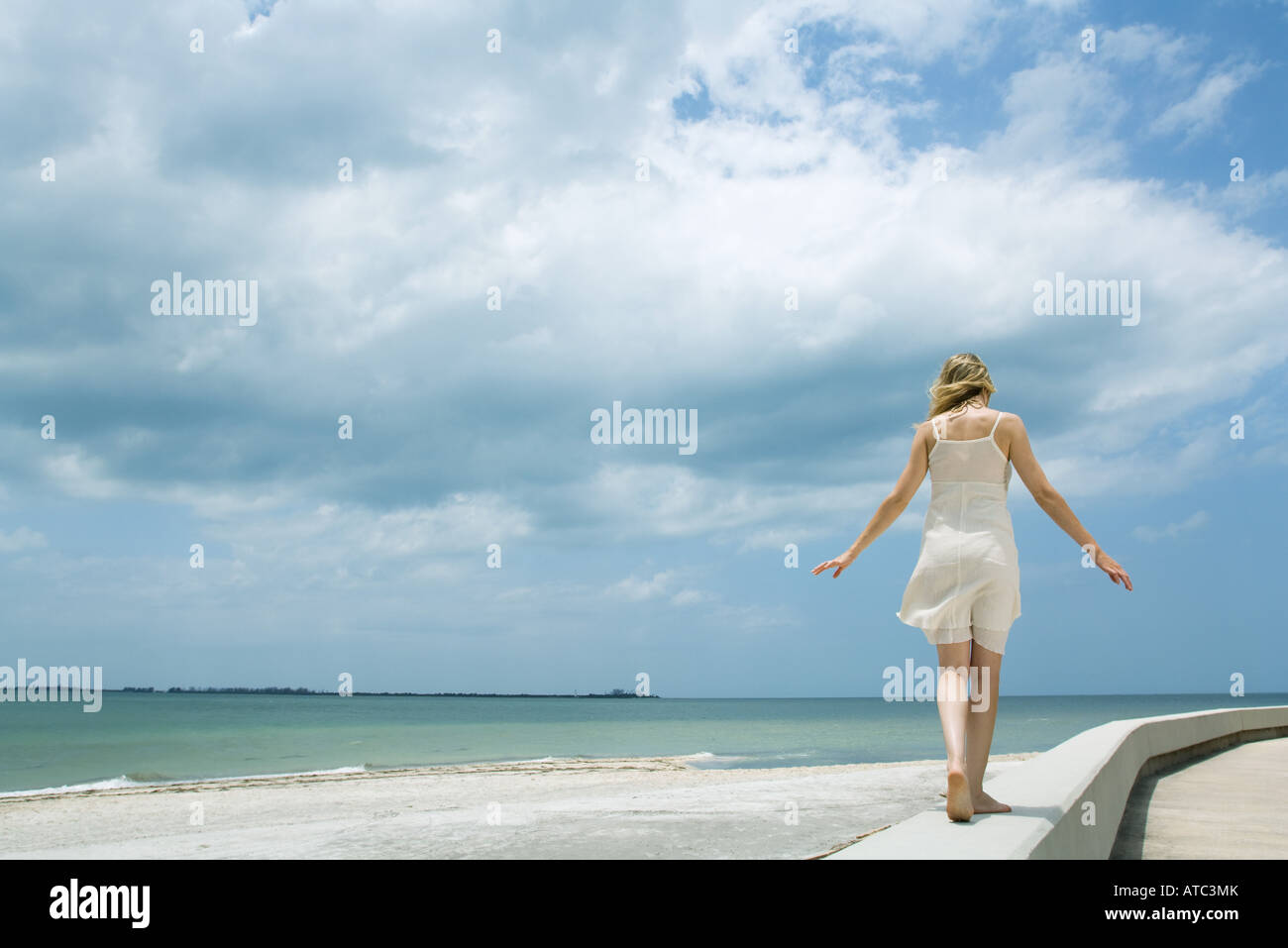 Young woman in sundress walking along low wall at the beach, arms out ...
