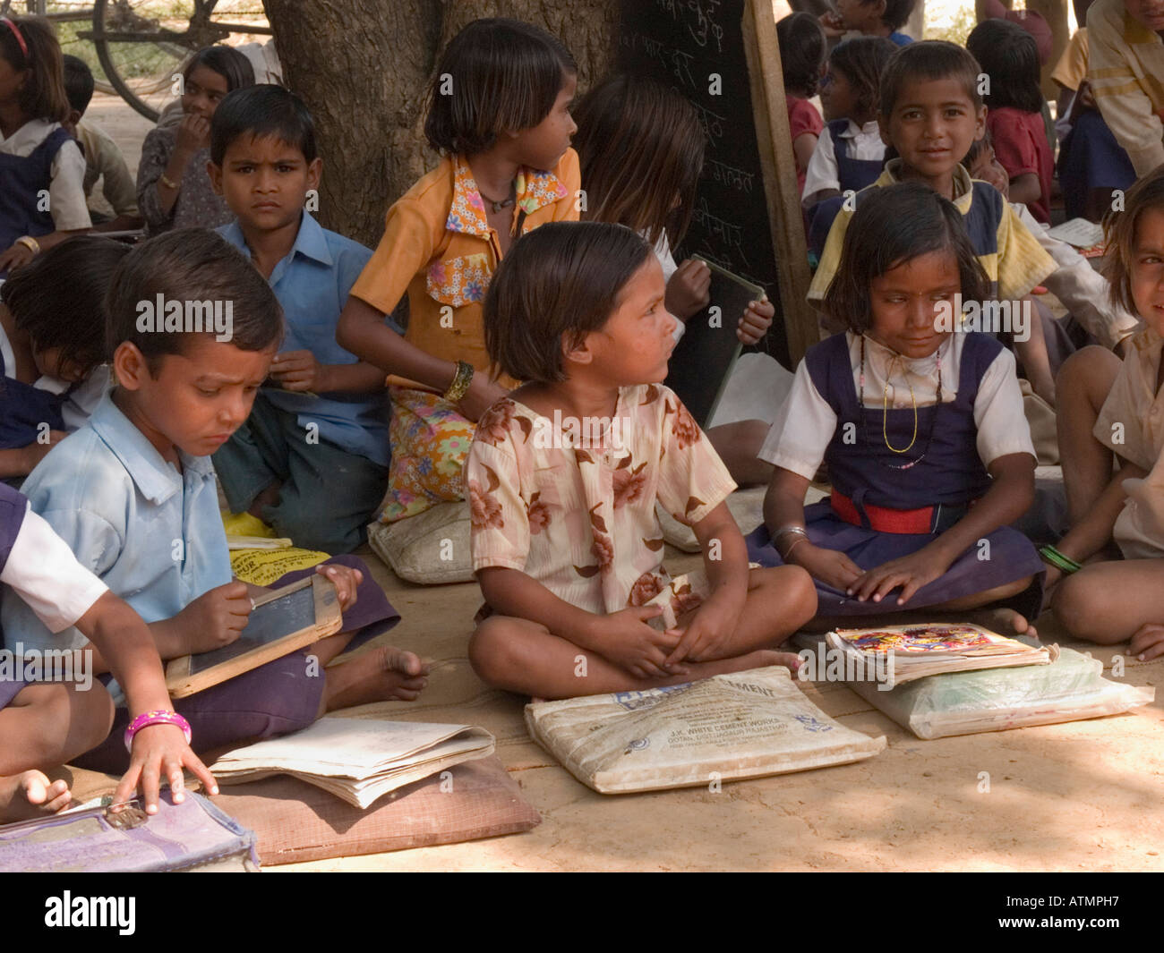 Junior school children sitting on ground with books in shade in rural ...