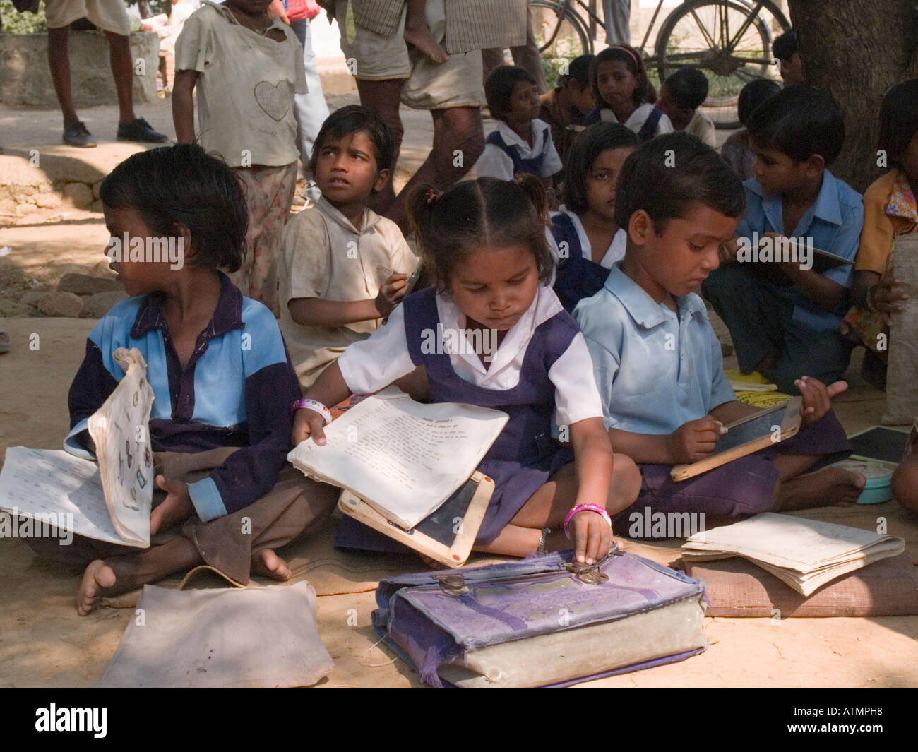 Junior school children sitting on ground with books in shade in rural ...