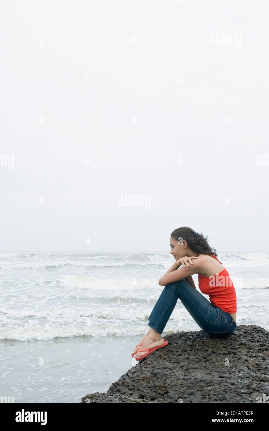 Side profile of a young woman sitting on a rock and looking at the sea ...