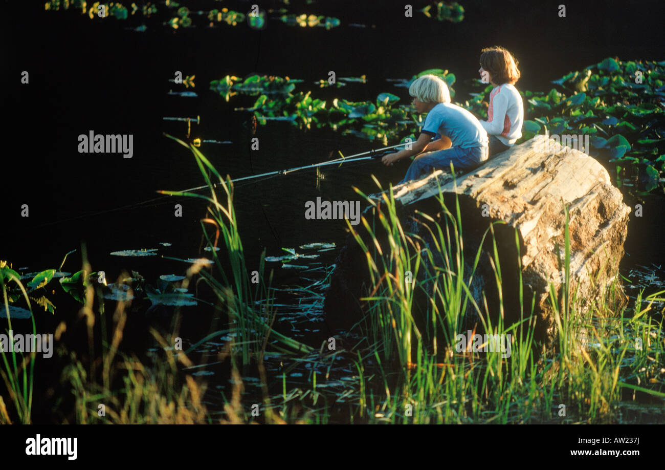 Boy and girl sitting on giant log next to pond fishing in sunset light ...