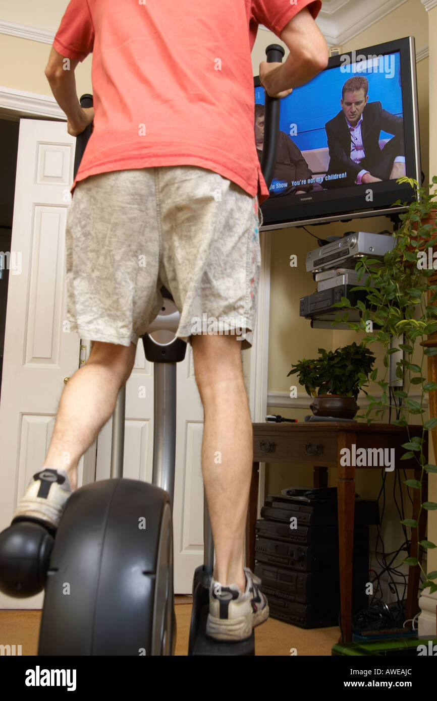 man on exercise machine at home changing TV channel with remote control ...