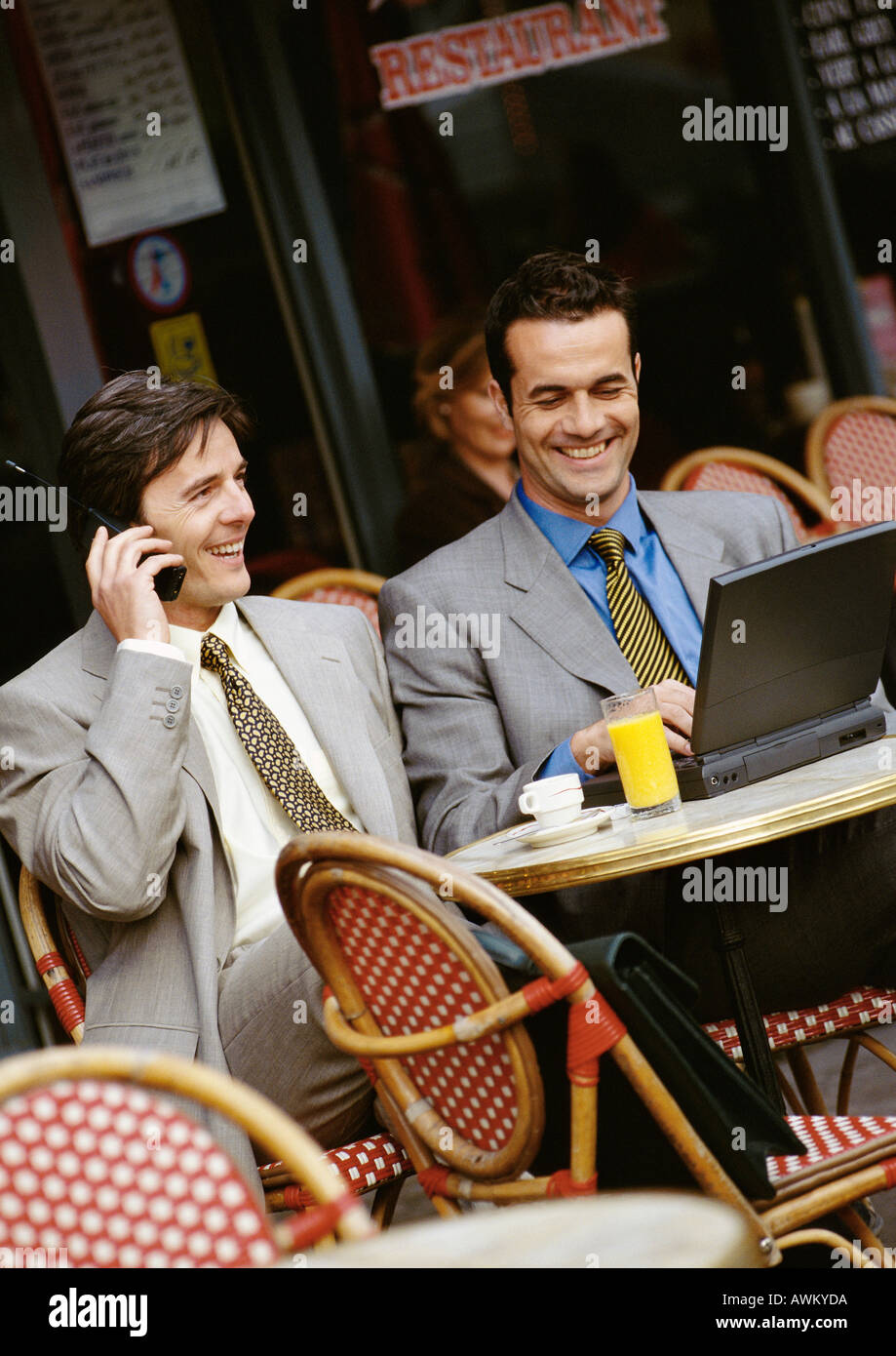 Two businessmen sitting at cafe terrace, using cell phone and laptop ...