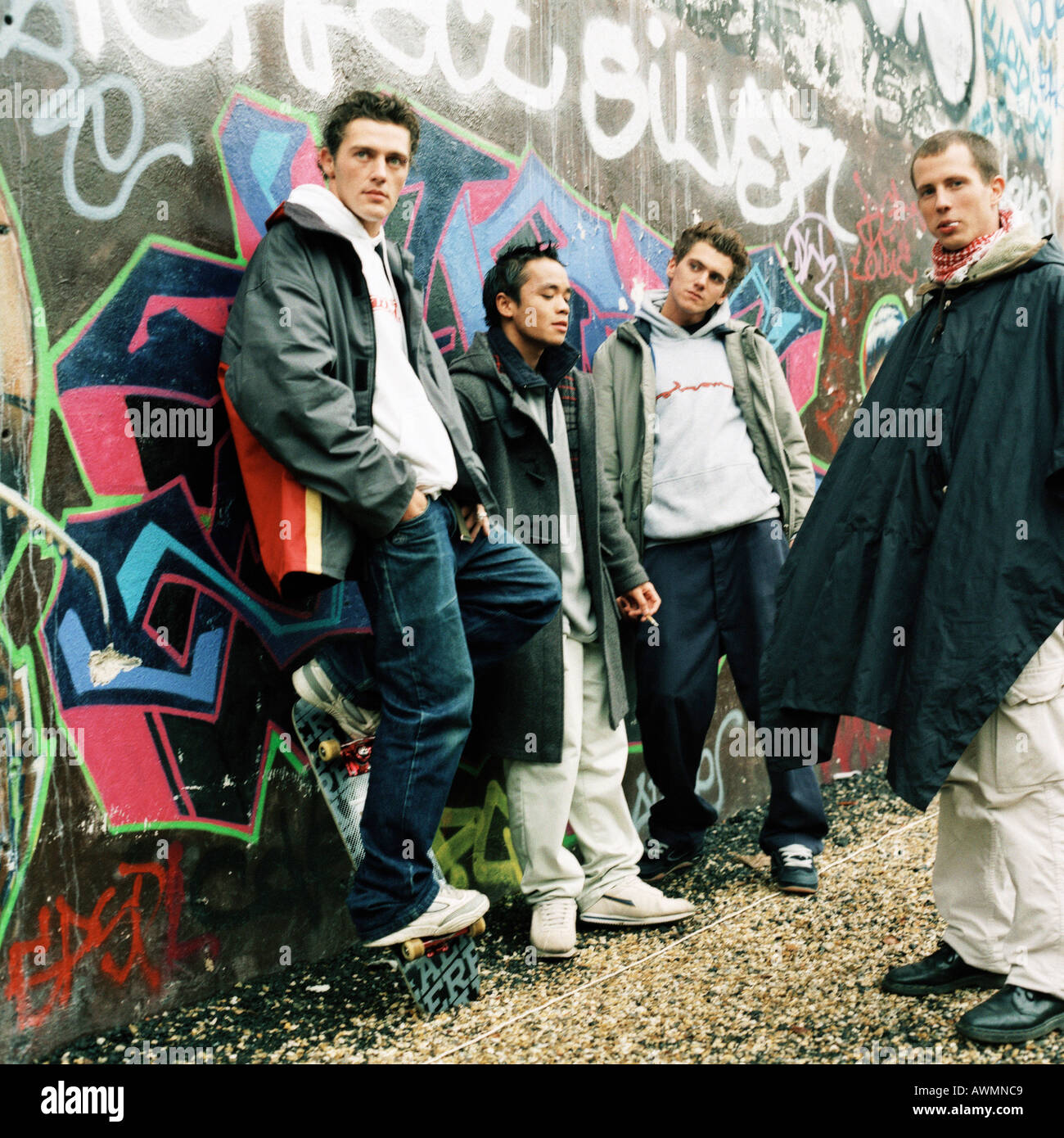 Four young men in city street, three leaning against graffiti wall ...
