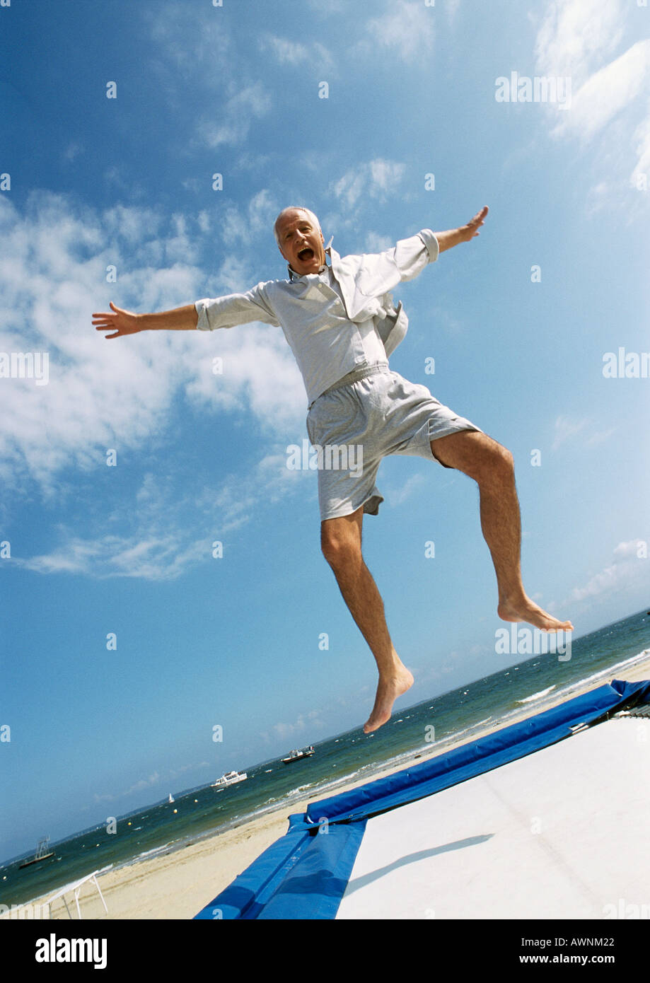 Mature man jumping on trampoline at the beach, arms out, mouth open ...