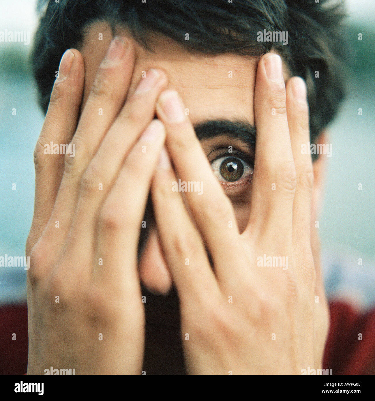 Young man with hands over face, looking through fingers with one eye ...