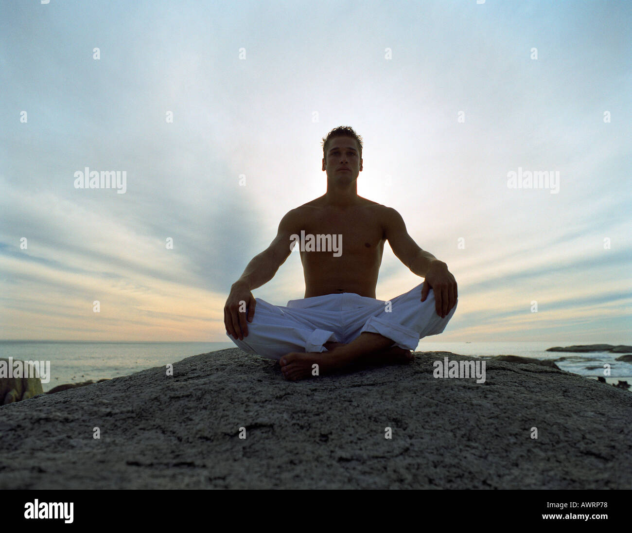 Man sitting indian style on rock, backlit, full length, sky and sea in ...