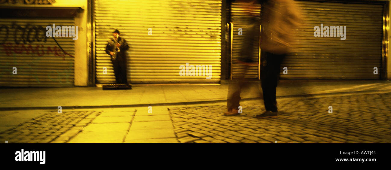 Musician leaning against roller shutter, people walking on cobble stone ...