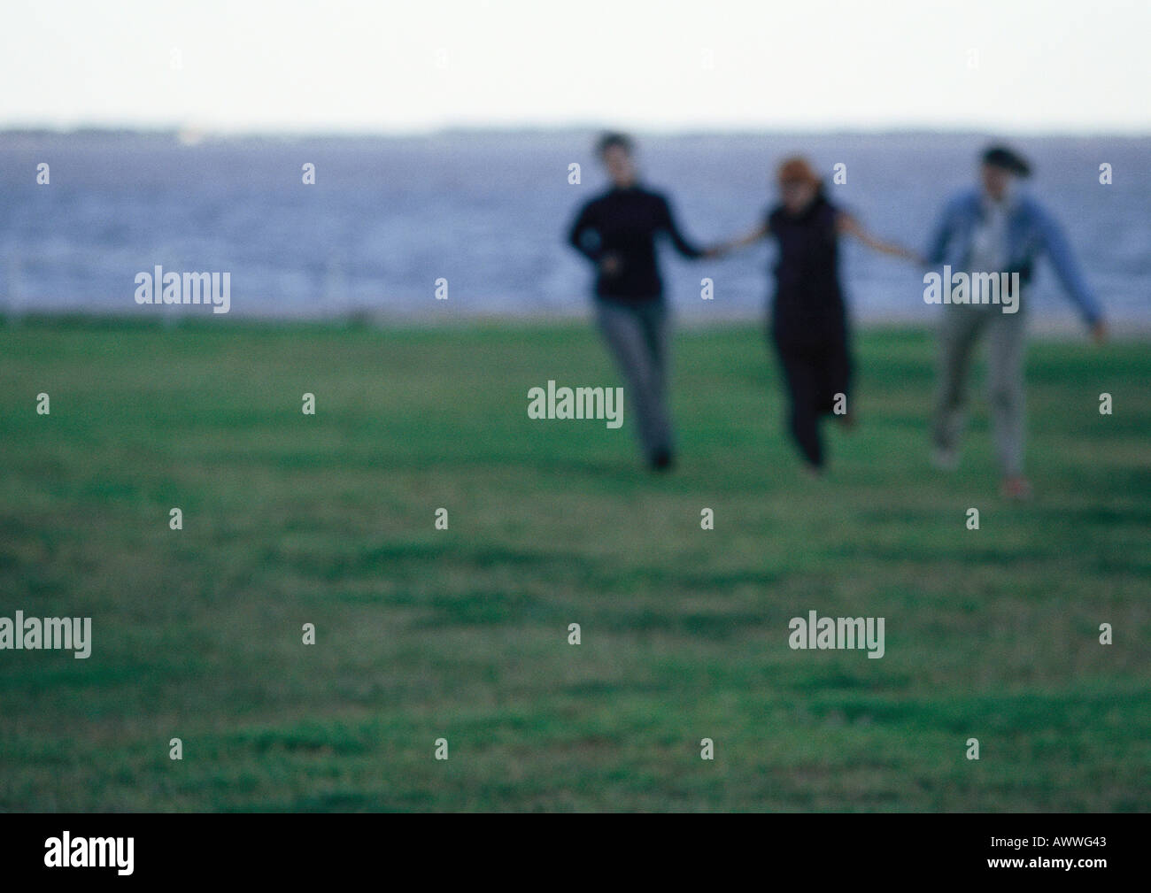 Three women, hand in hand, running across field of grass, sea on ...