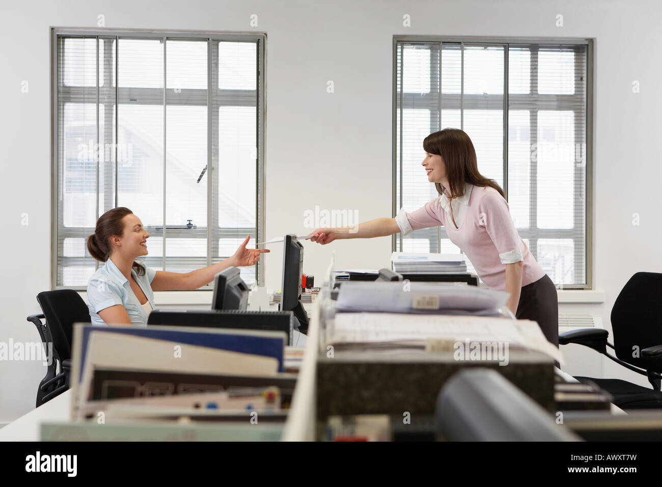Two women in office, one handing documents to other over desk, side ...