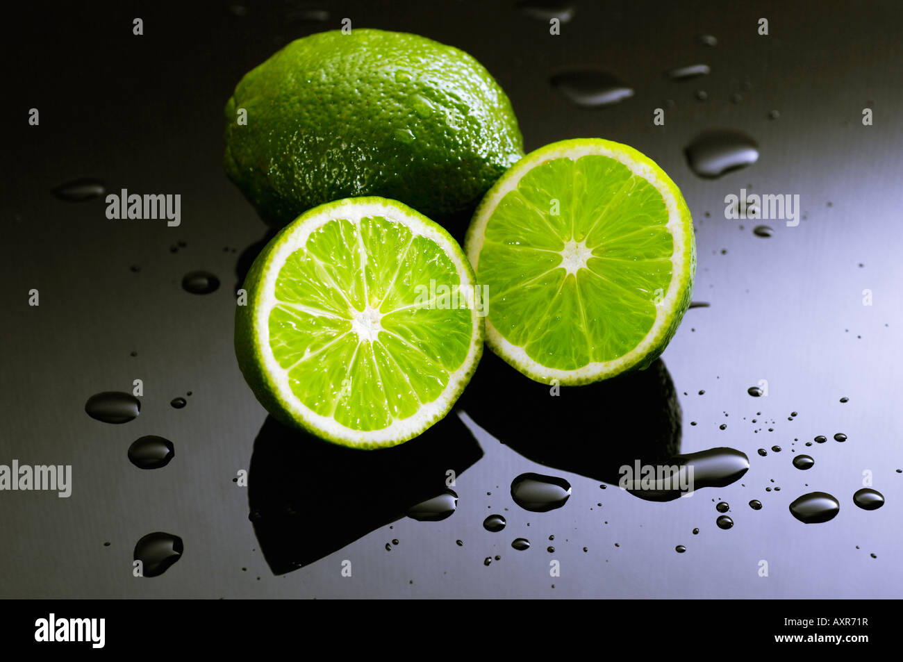Fresh limes, one cut in half and one whole on a black reflective ...