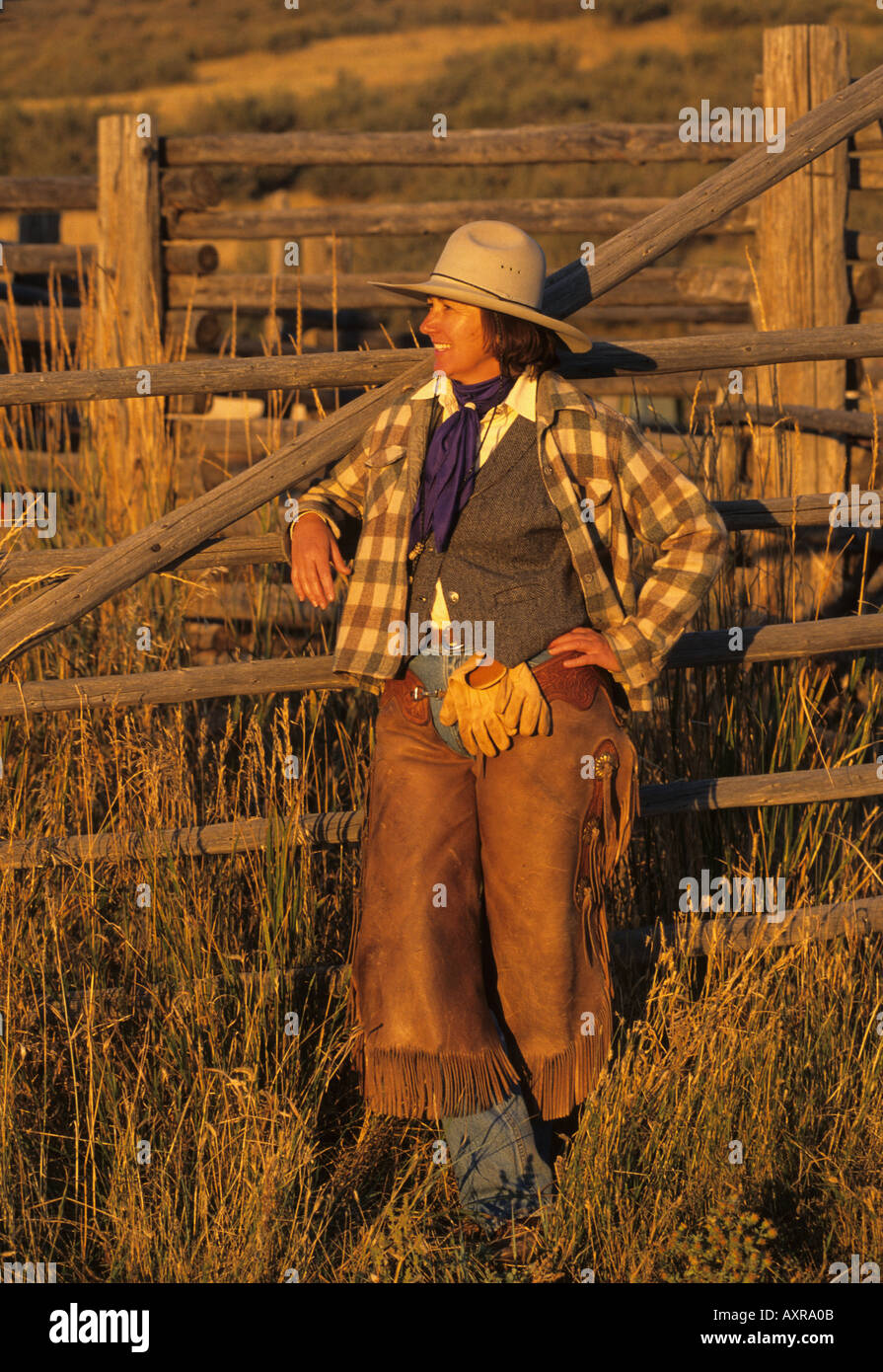 Smiling female rancher in chaps and cowboy hat stands by corrals adn in ...