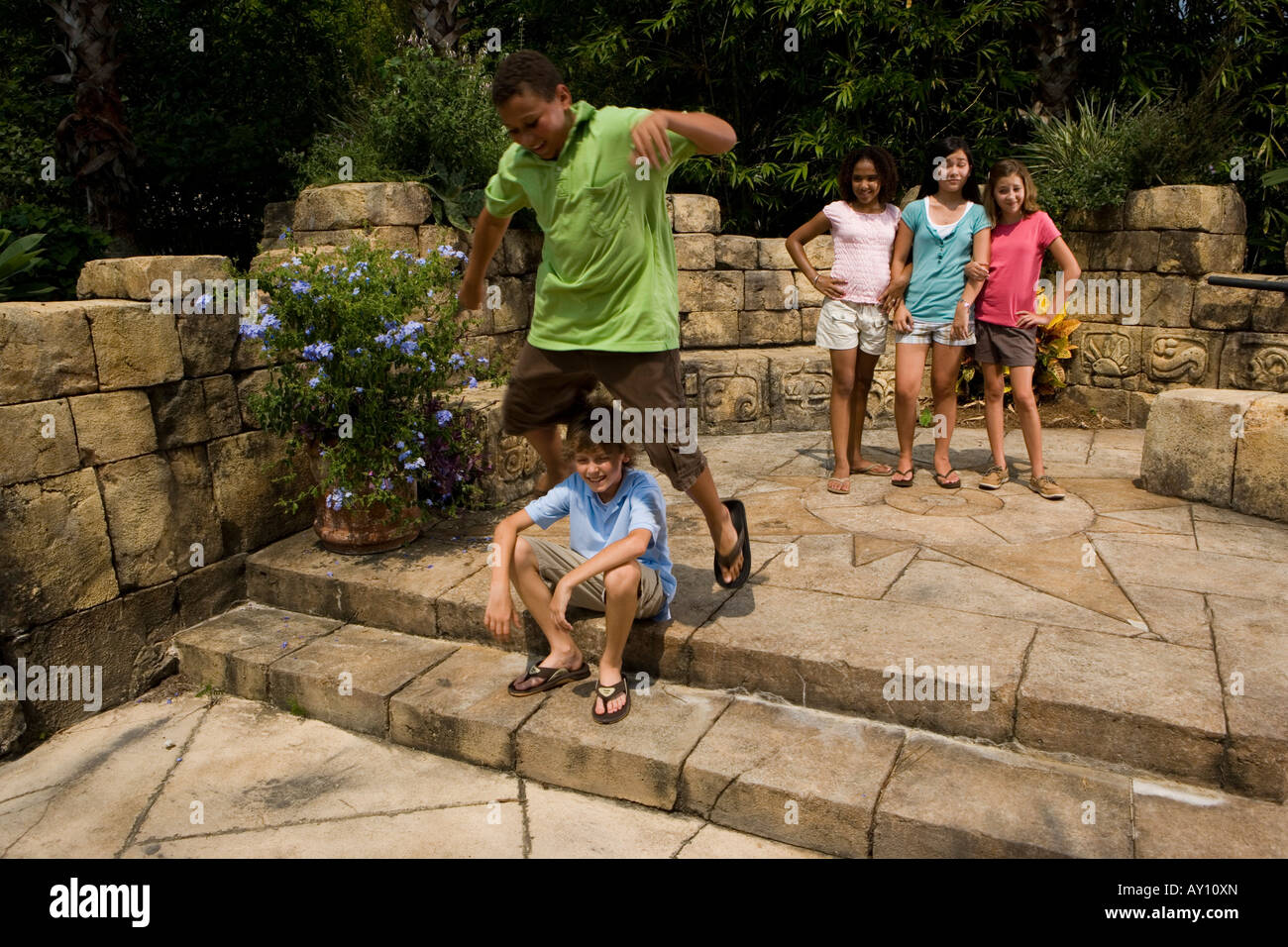 Teenage boy jumping over his friend with girls standing in background ...