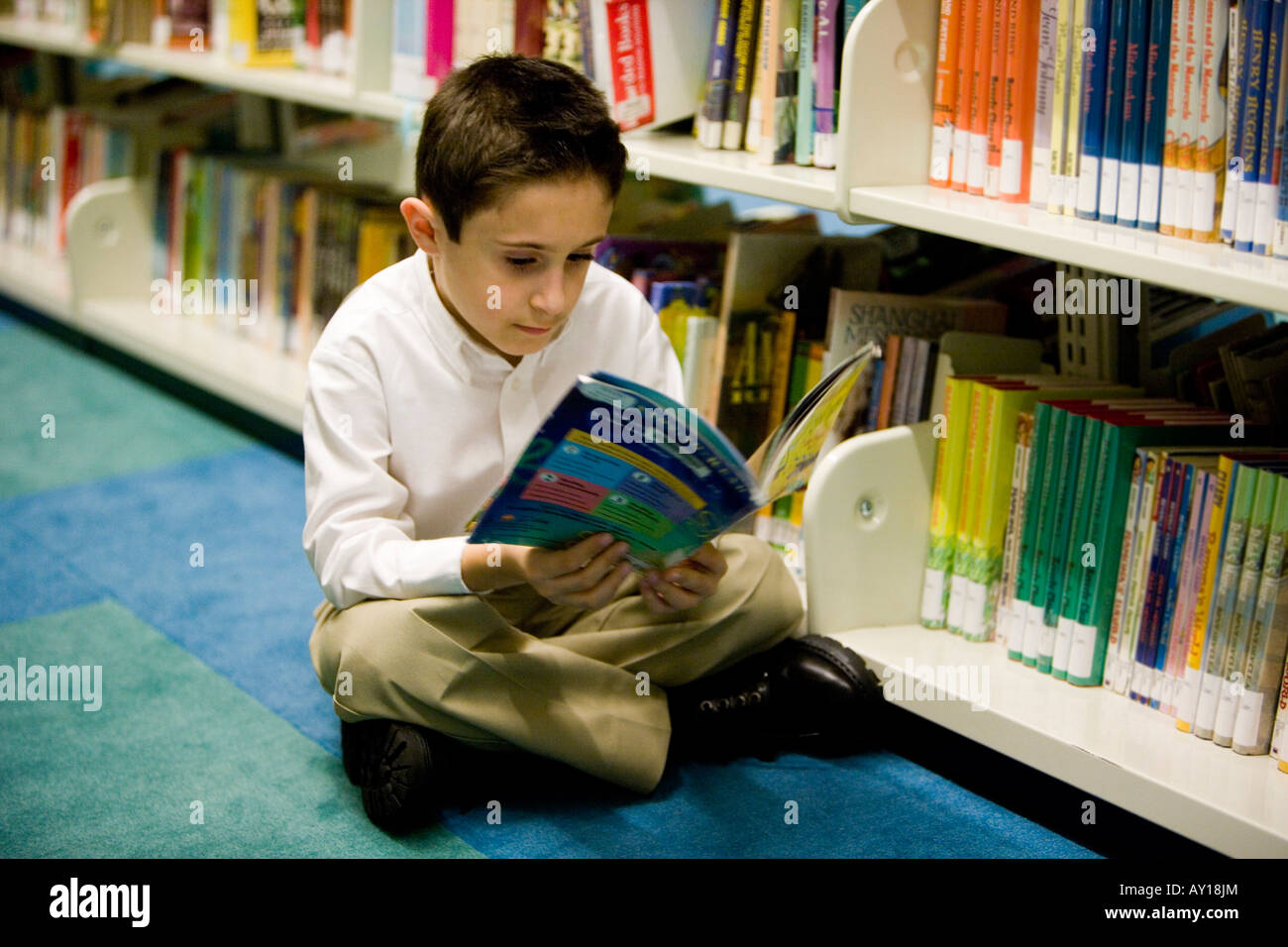 Schoolboy sitting on carpet and reading a book in the library Stock ...