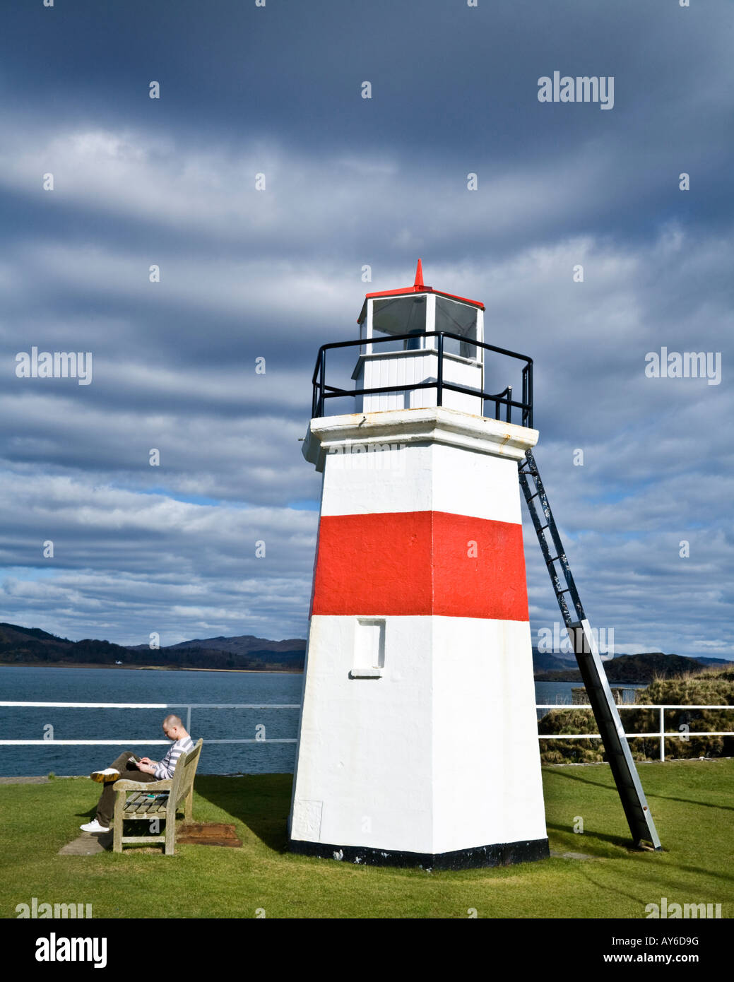 A man sitting on a bench reading by the Crinan canal harbour lighthouse ...