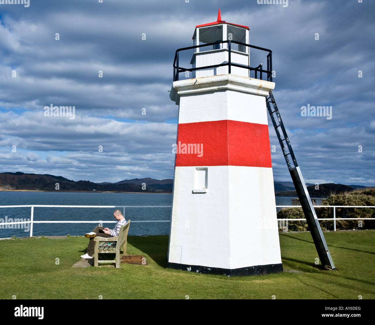 A man sitting reading on a bench by the Crinan canal harbour lighthouse ...
