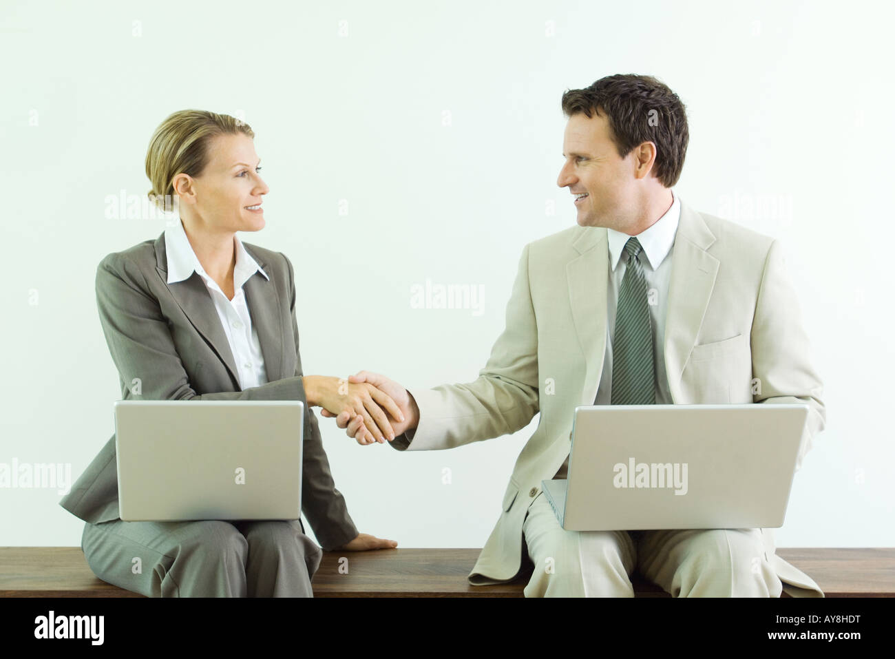 Two business associates sitting, shaking hands, both using laptop ...