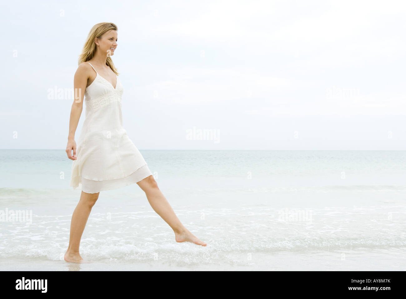 Young woman in sundress taking large step as she walks in shallow water ...