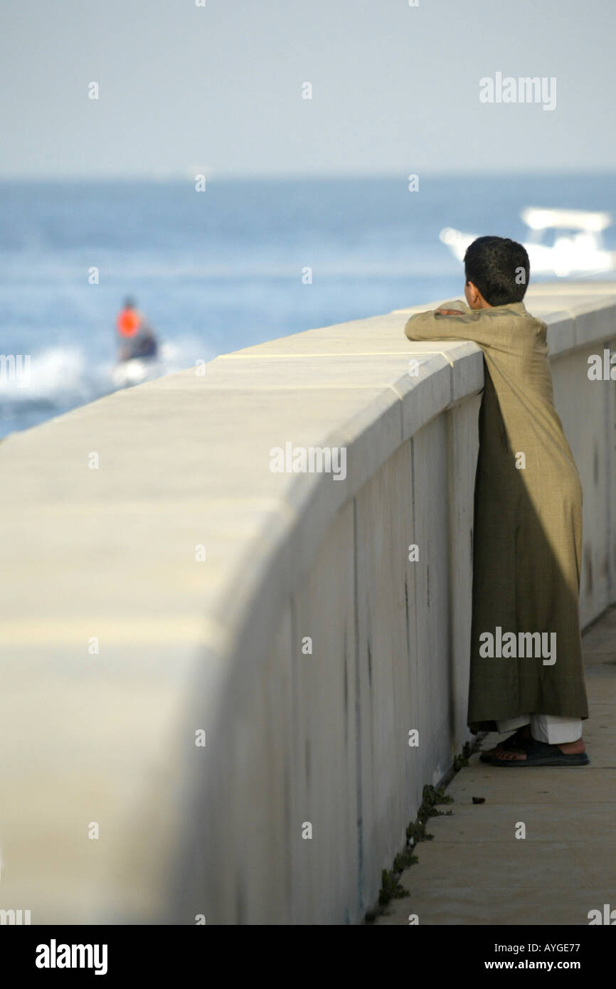A young Kuwaiti boy in a traditional thaub watches people ride jet skis ...