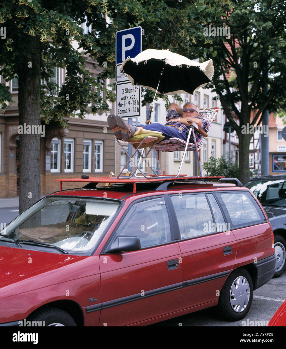 man lies in a deckchair with a sunshade on top of his car, parking lot ...