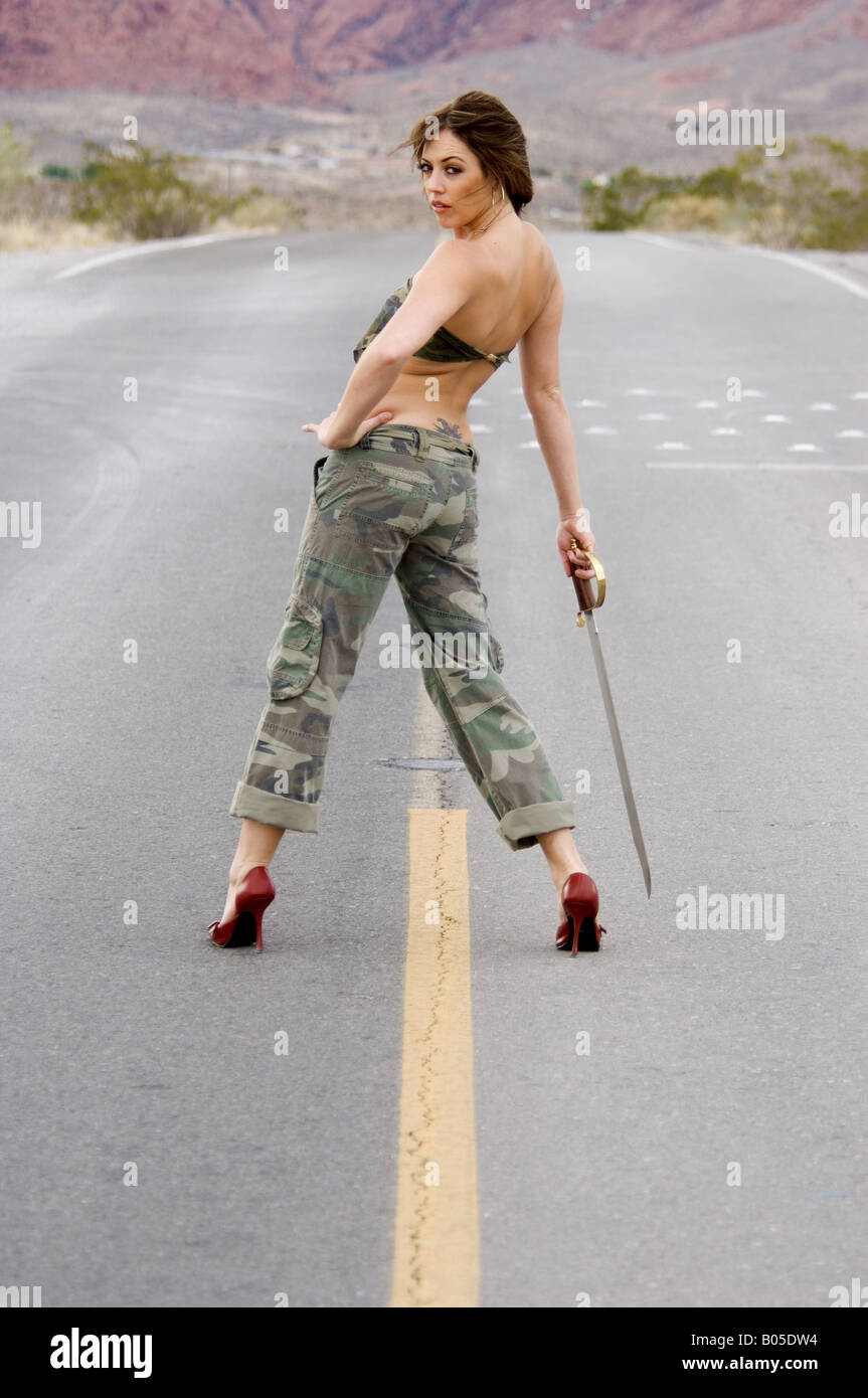 woman with a sword in fatigues on a country road in the desert looking ...