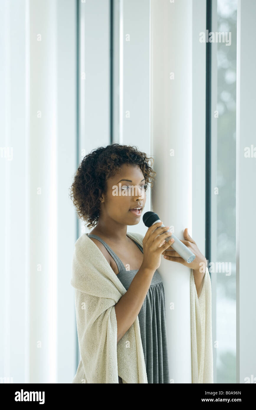 Young woman leaning against pole, singing into microphone, looking away ...