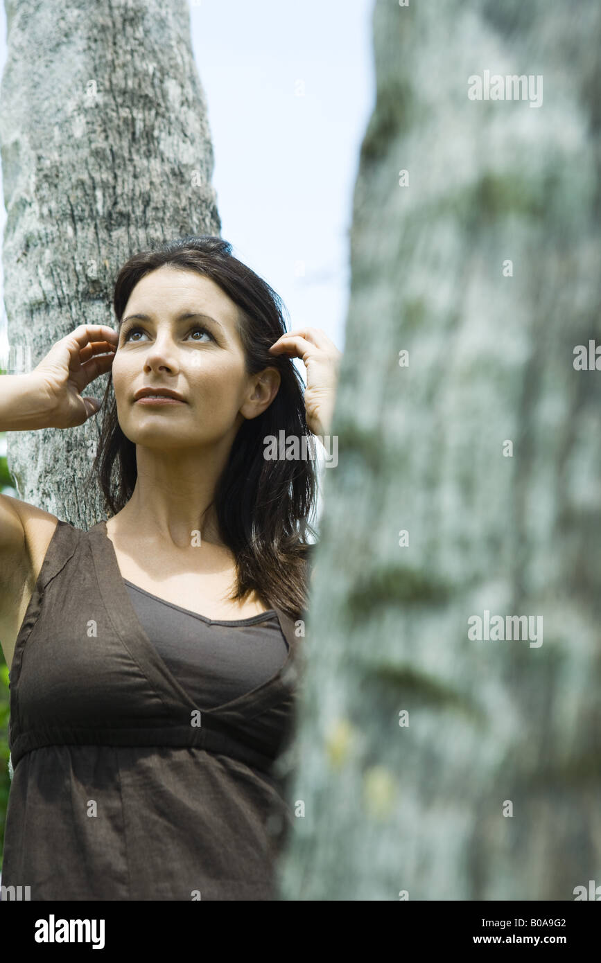 Woman leaning against tree trunk, pushing hair behind ears, looking up ...