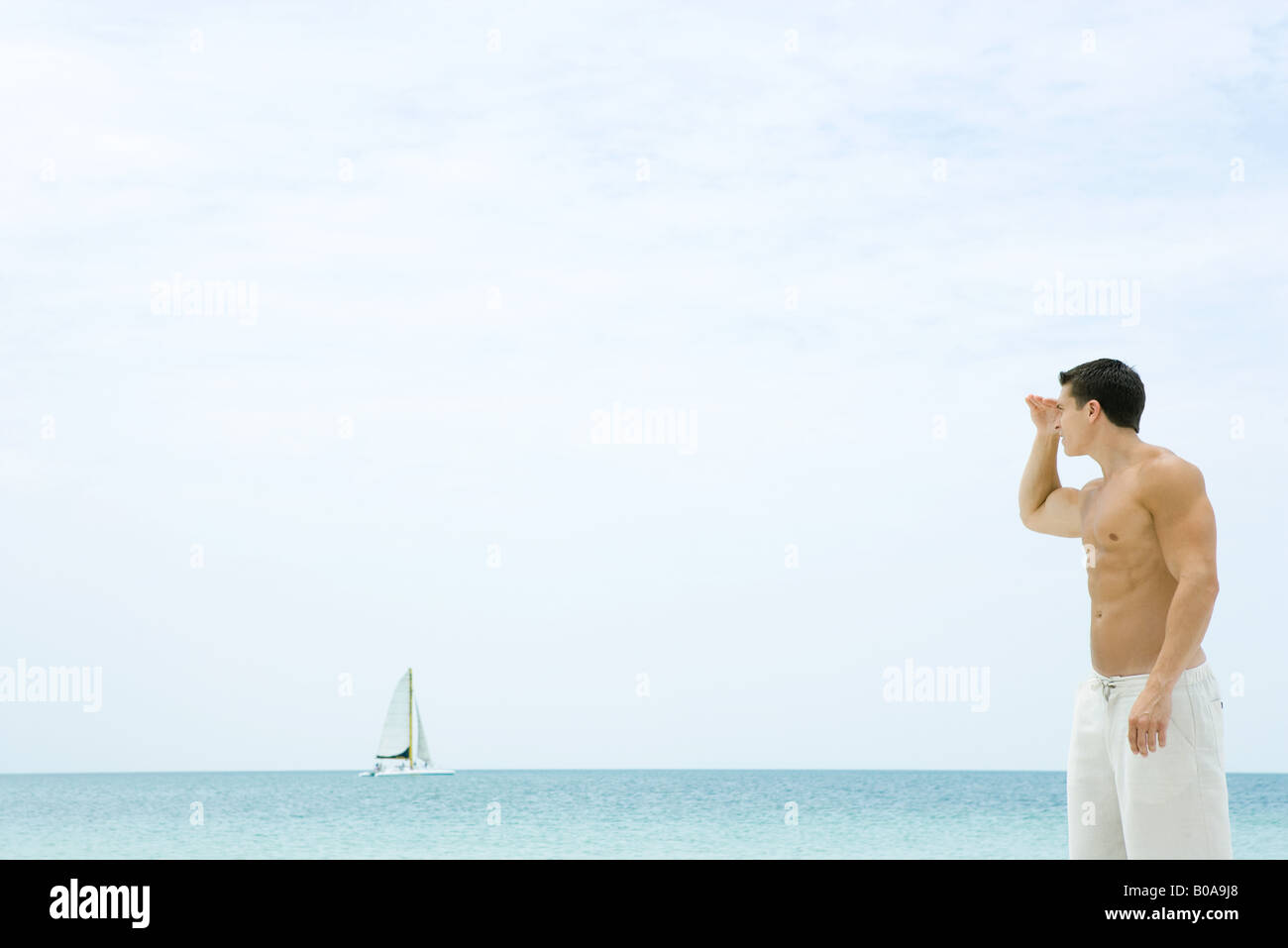 Man standing at the beach, looking at view, sailboat in the distance ...