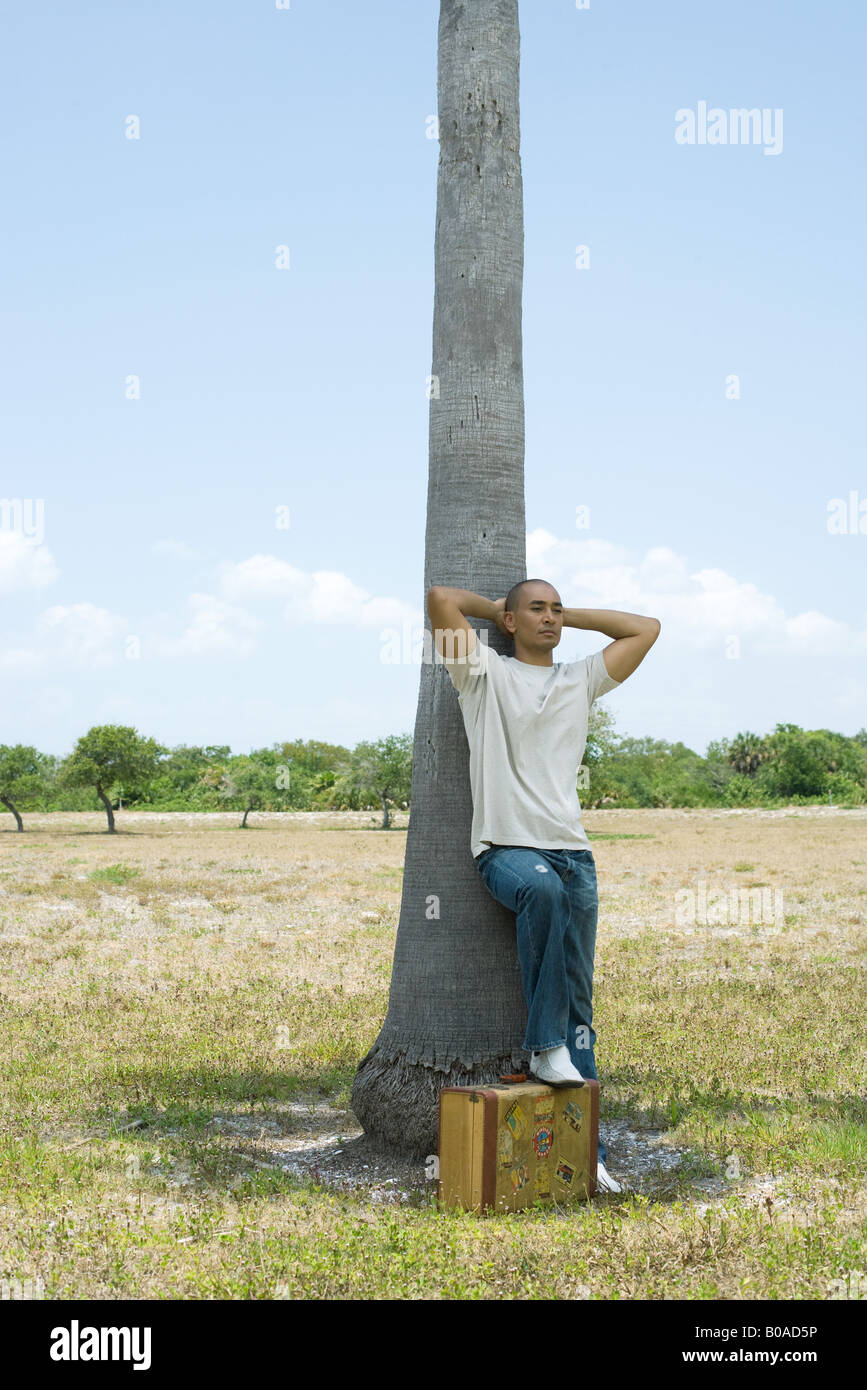 Man leaning against tree trunk, hands behind head, one foot resting on ...