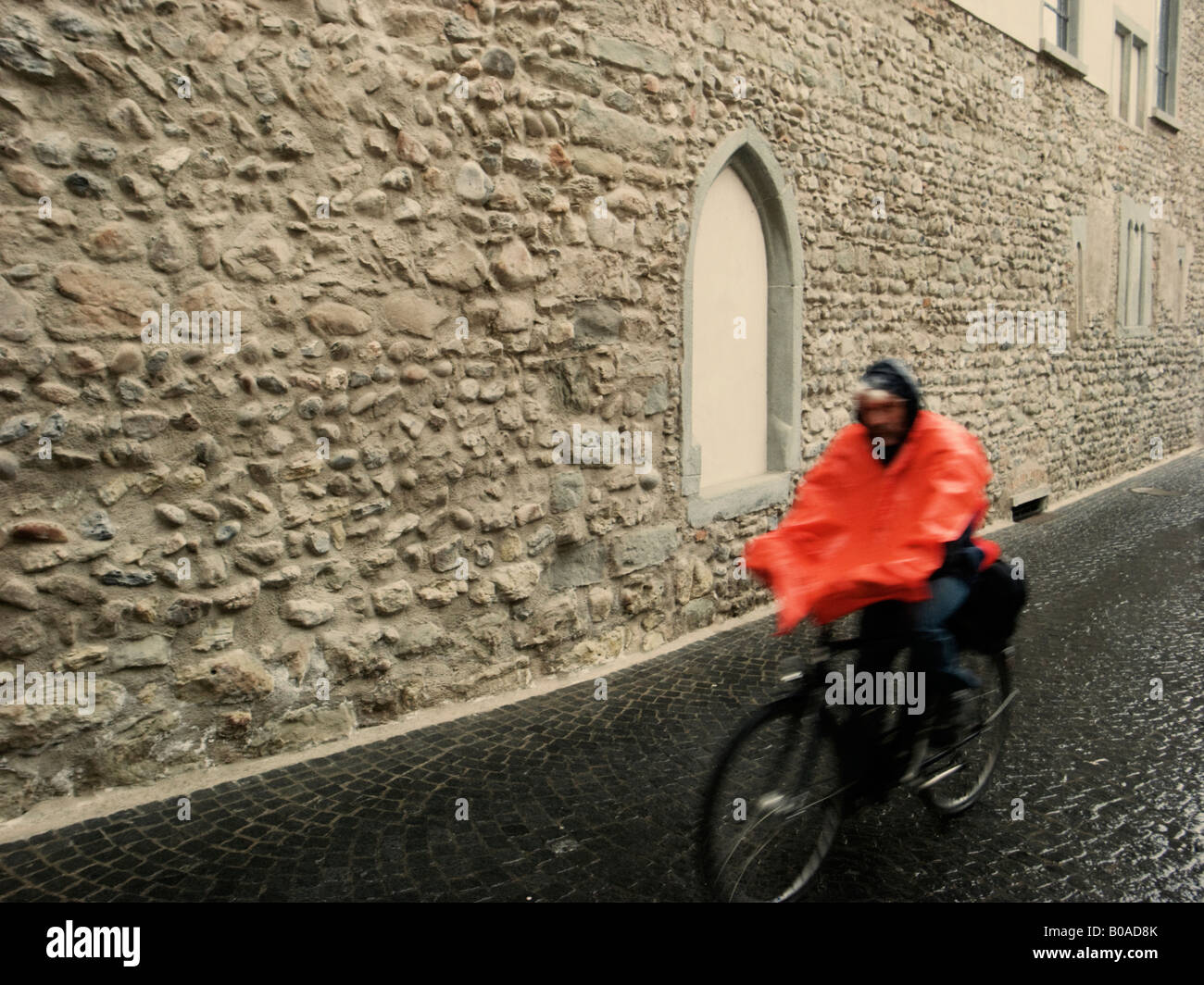 Postman on a bicycle,cycling in the rain on the Cobbled street of the ...