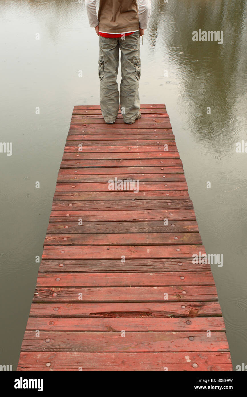 Teenage boy standing at the end of a wooden pier jetty on a lake as it ...