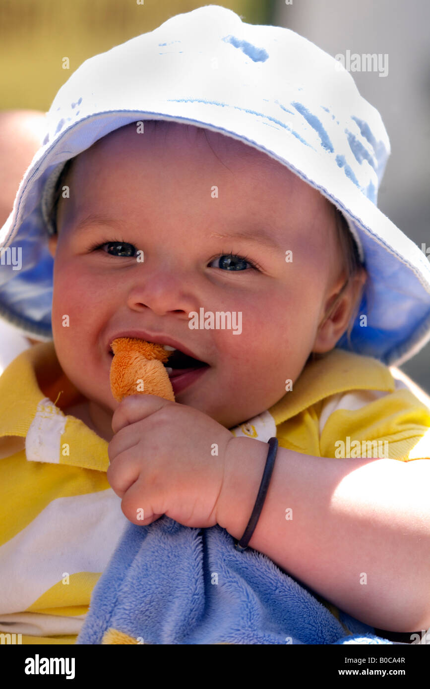 Toddler outdoors wearing blue hat, putting soft toy in his mouth ...