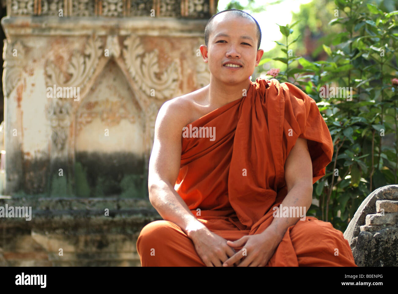 A Buddhist monk dressed in bright orange robes sits on a wall at a ...