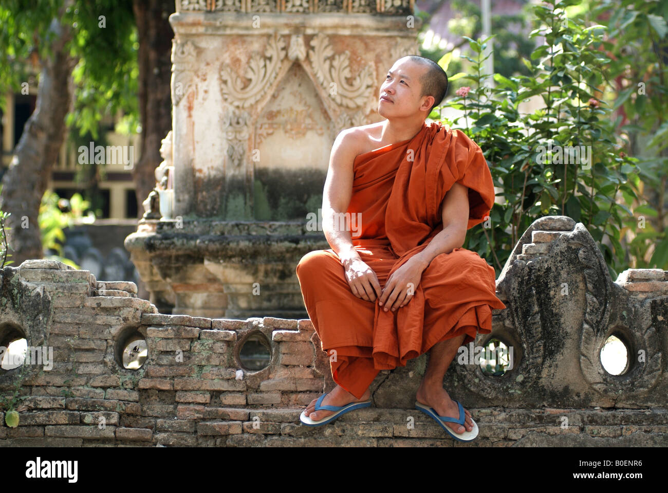 A Buddhist monk dressed in bright orange robes sits on a wall at a ...
