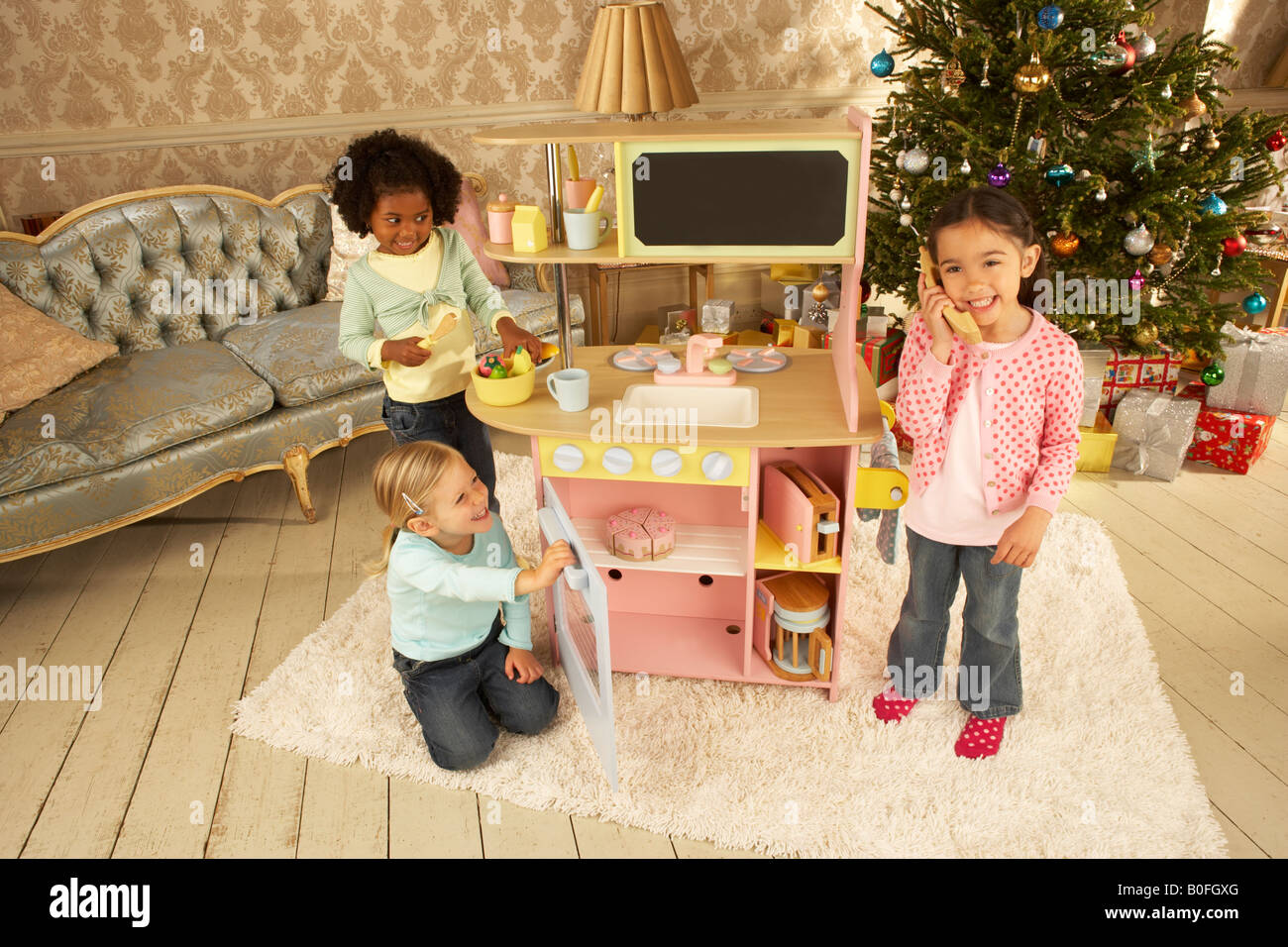 three girls play at Christmas time under the tree with a toy kitchen ...