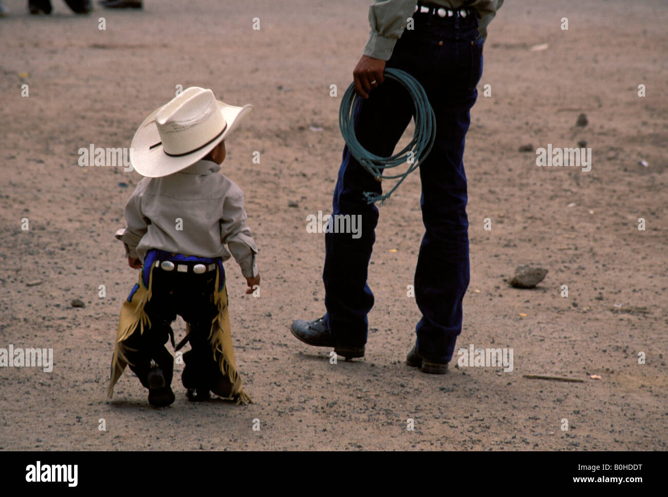 A young boy dressed as a cowboy at a Navajo pow-wow following an adult ...