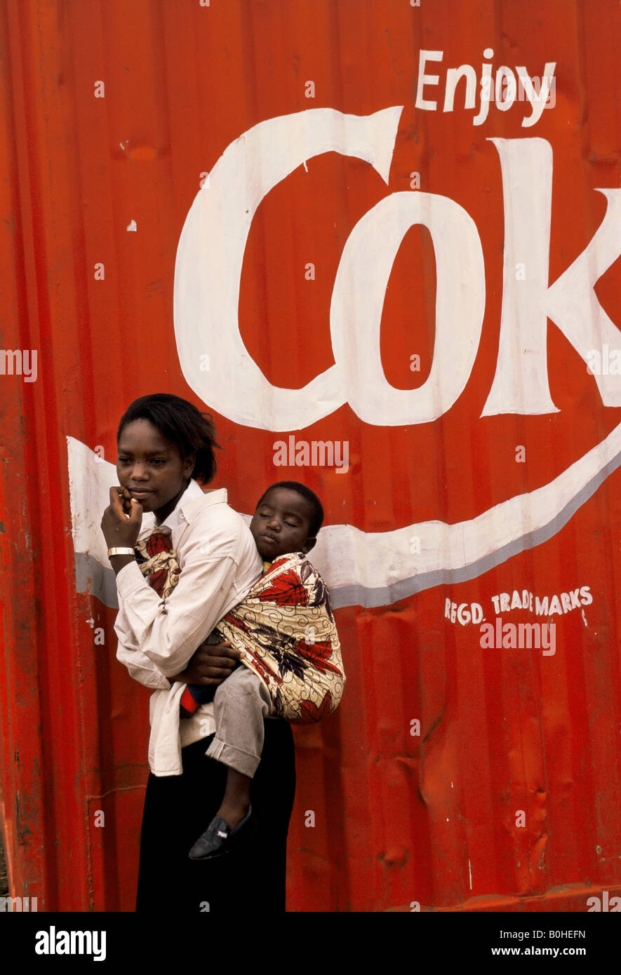 A girl carrying a baby in front of a Coca-Cola advertisement, Lusaka ...