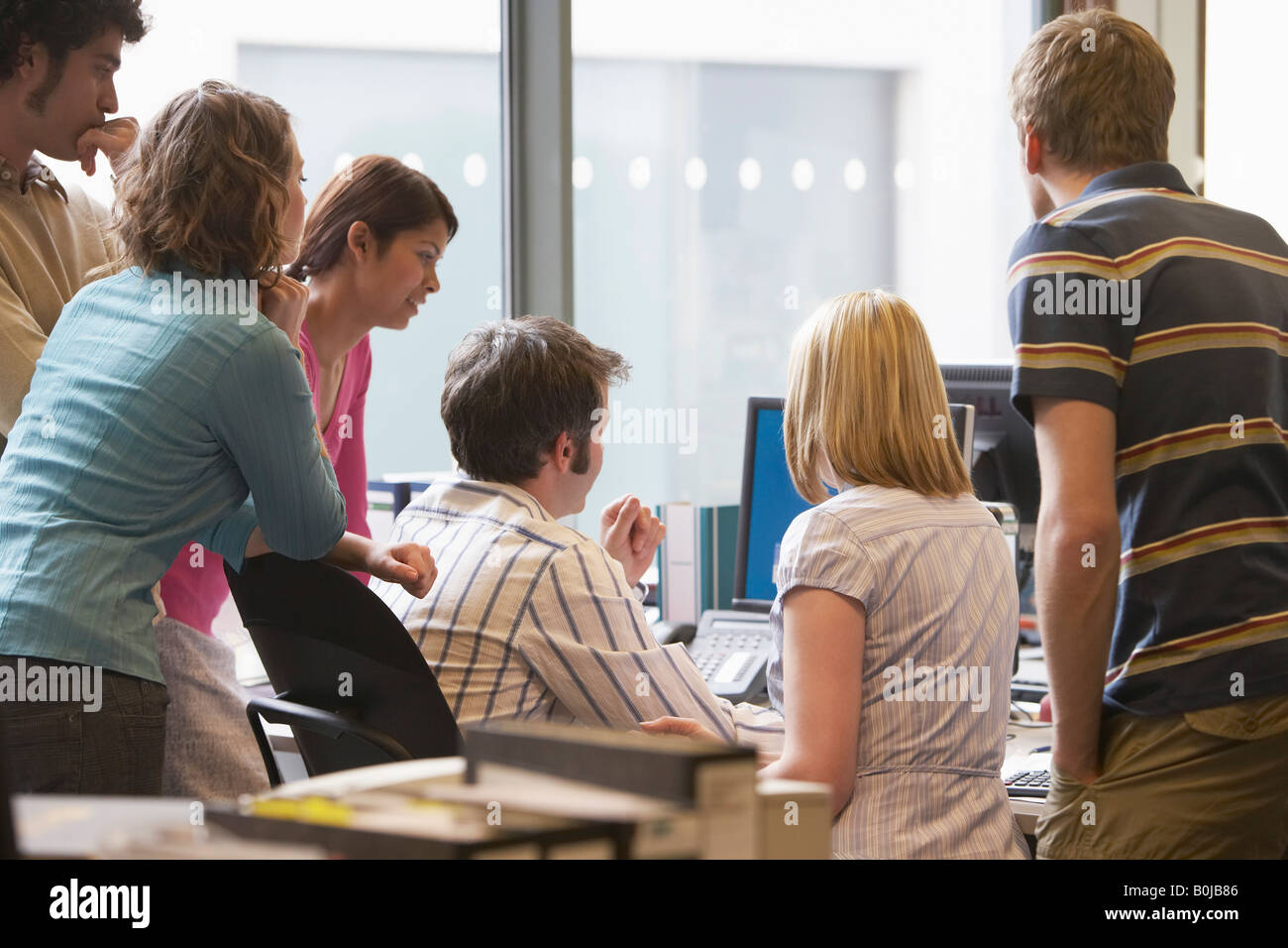 Office workers standing around colleague using computer, back view ...